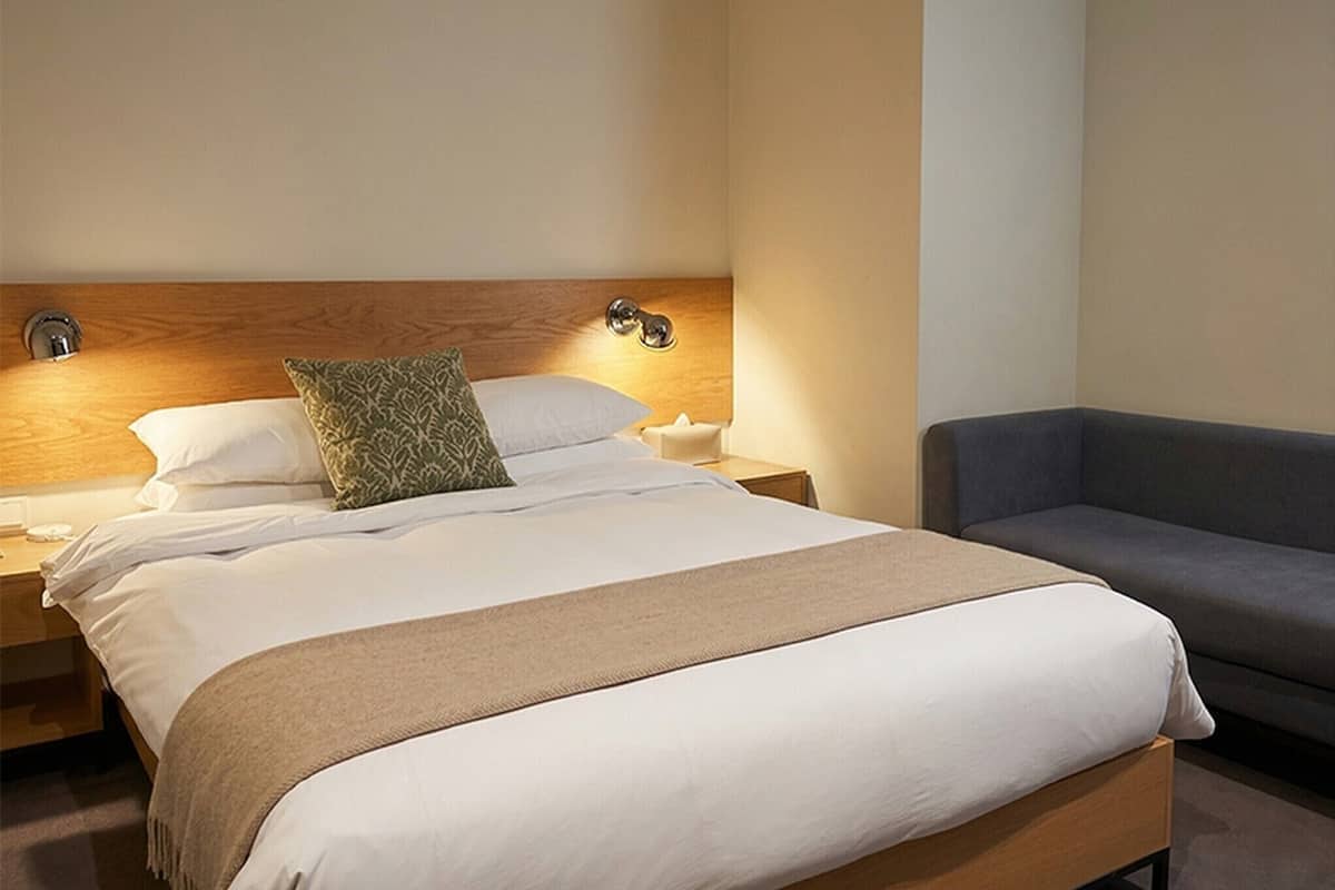 Quarto de hotel com cama, mesa de cabeceira, sofá e luminária fixada na parede.