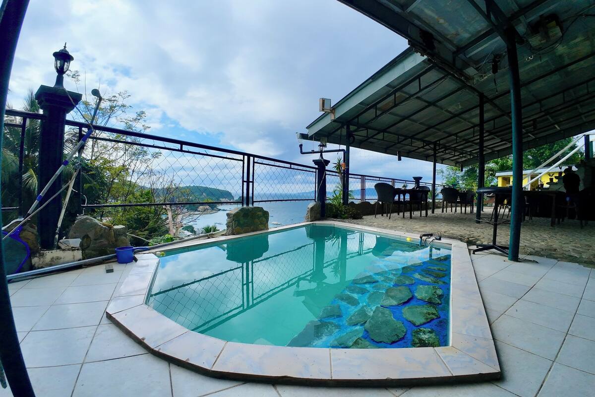 Piscina com vista para o mar, rodeada por um terraço coberto com cadeiras e mesas.