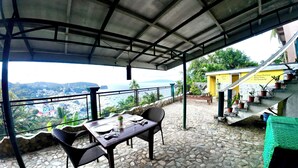 Uma área de estar coberta ao ar livre, com mesa e cadeiras, oferece vista panorâmica de uma cidade costeira e montanhas.