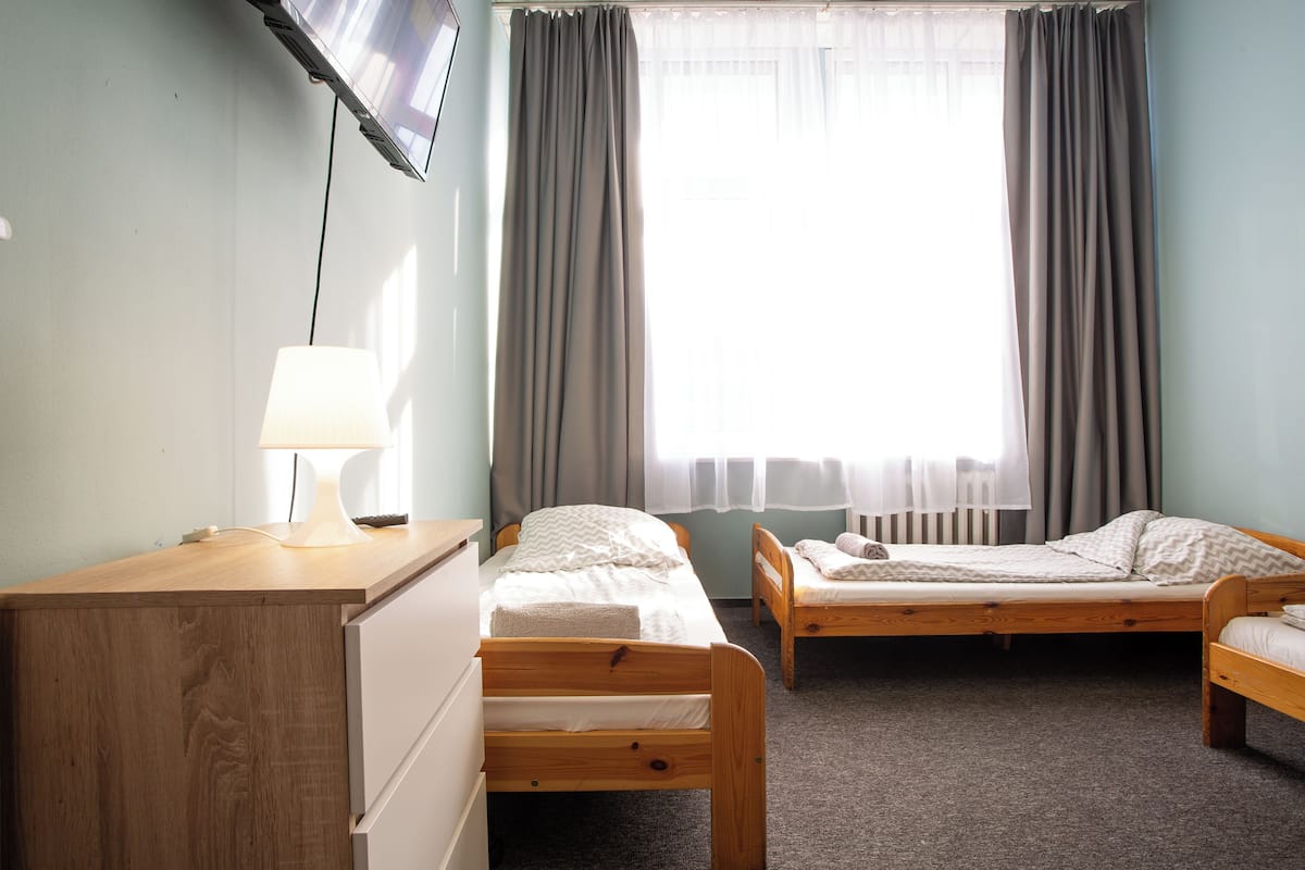 Quarto com duas camas de madeira, uma escrivaninha com abajur, um radiador e uma TV fixada na parede.