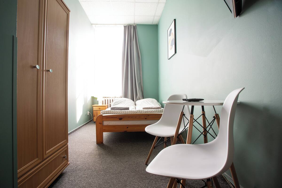 Quarto com cama, escrivaninha, duas cadeiras, guarda-roupa e janela com cortinas.