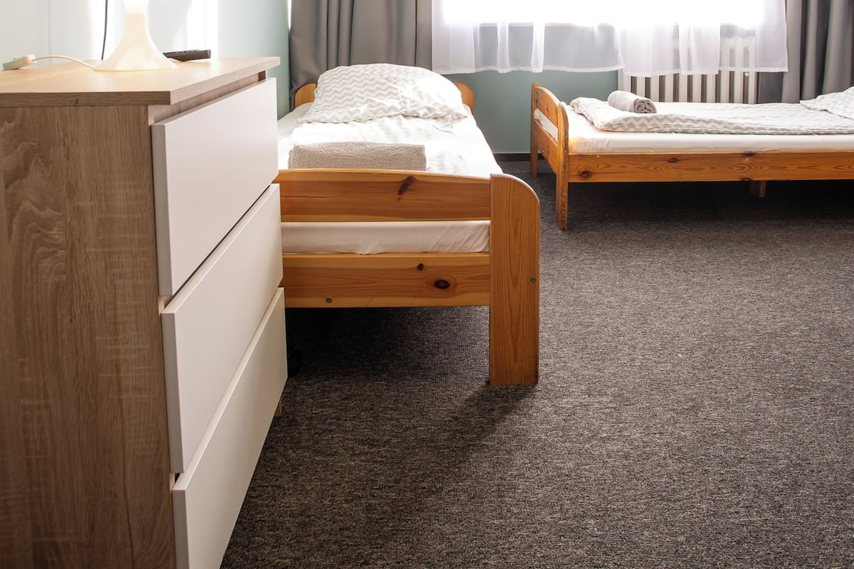 Quarto com duas camas de madeira, uma escrivaninha com luminária, televisão na parede e uma janela com cortinas.