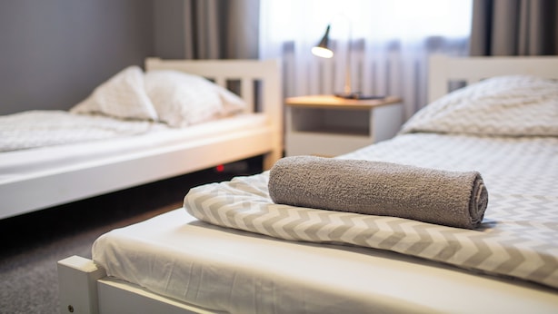 Duas camas de solteiro com roupa de cama branca e uma toalha enrolada, uma mesa de cabeceira com abajur e uma janela com cortinas.
