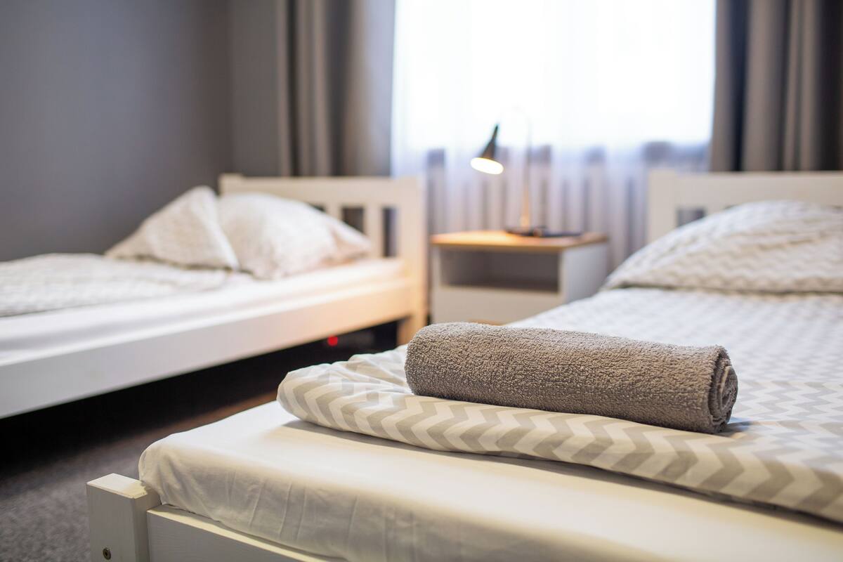 Duas camas de solteiro com roupa de cama branca e uma toalha enrolada, uma mesa de cabeceira com abajur e uma janela com cortinas.