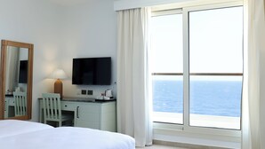 Quarto de hotel com cama, escrivaninha com luminária, televisão e uma grande janela com vista para o mar.