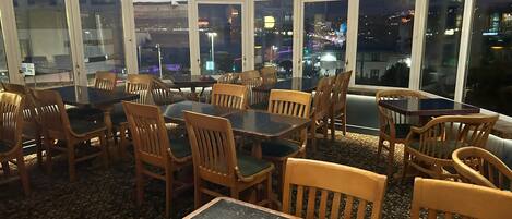 Um restaurante com várias mesas e cadeiras, grandes janelas oferecendo vista noturna da cidade.