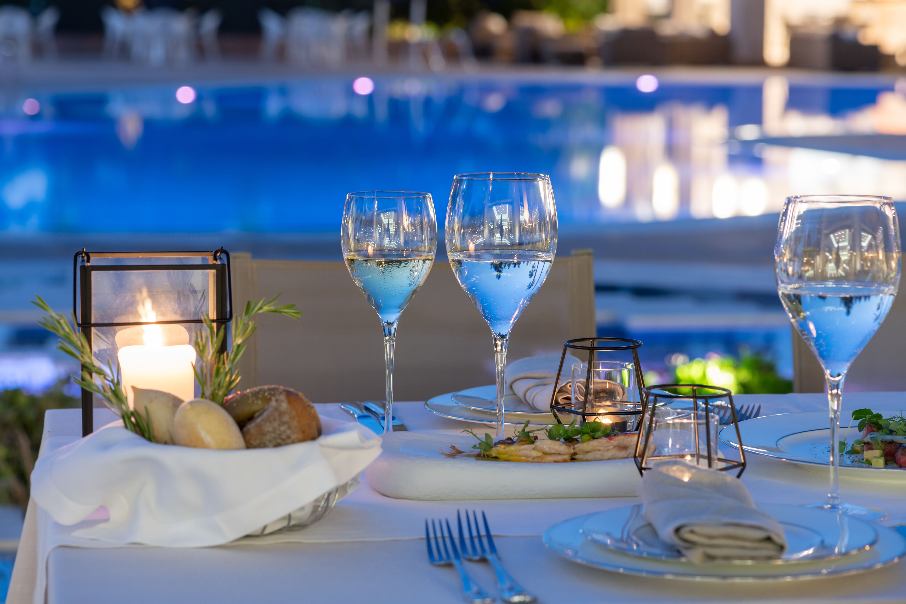 Mesa de jantar posta com velas, copos e um prato de comida, com vista para uma piscina.