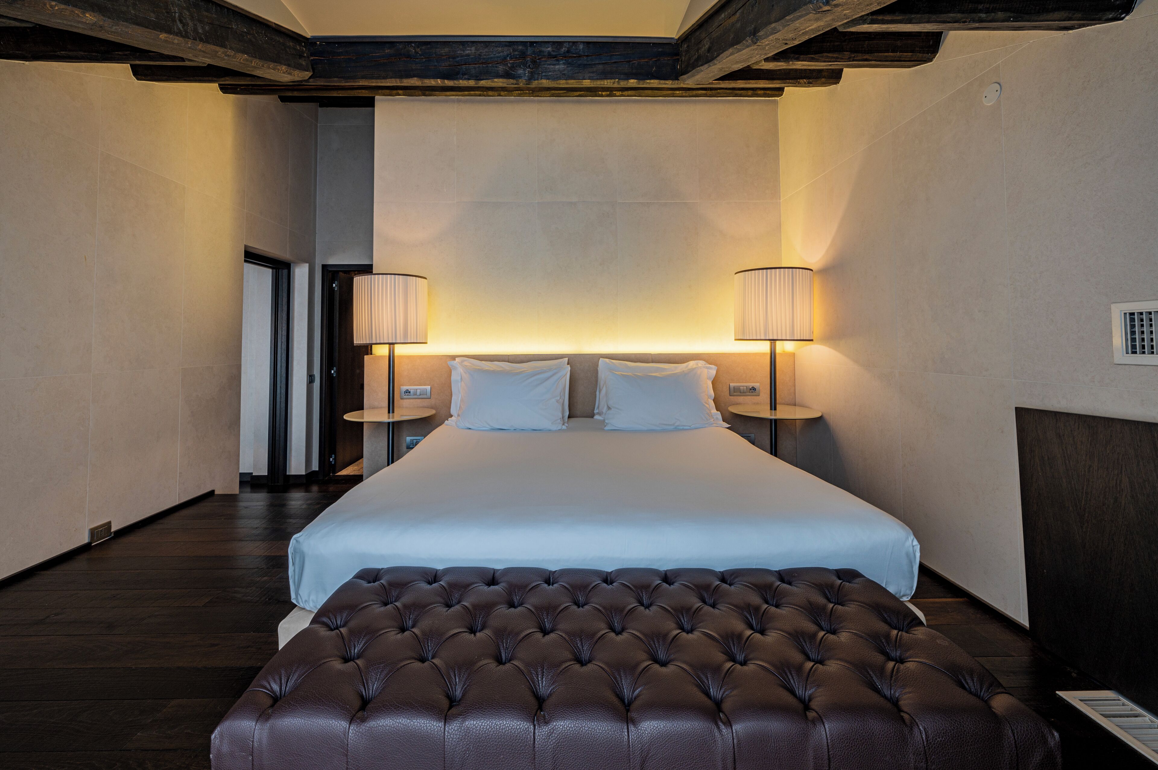 Quarto de hotel com cama grande, duas luminárias de cabeceira, um banco estofado marrom escuro e um vigamento de madeira no teto.