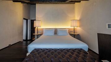 Quarto de hotel com cama grande, duas luminárias de cabeceira, um banco estofado marrom escuro e um vigamento de madeira no teto.