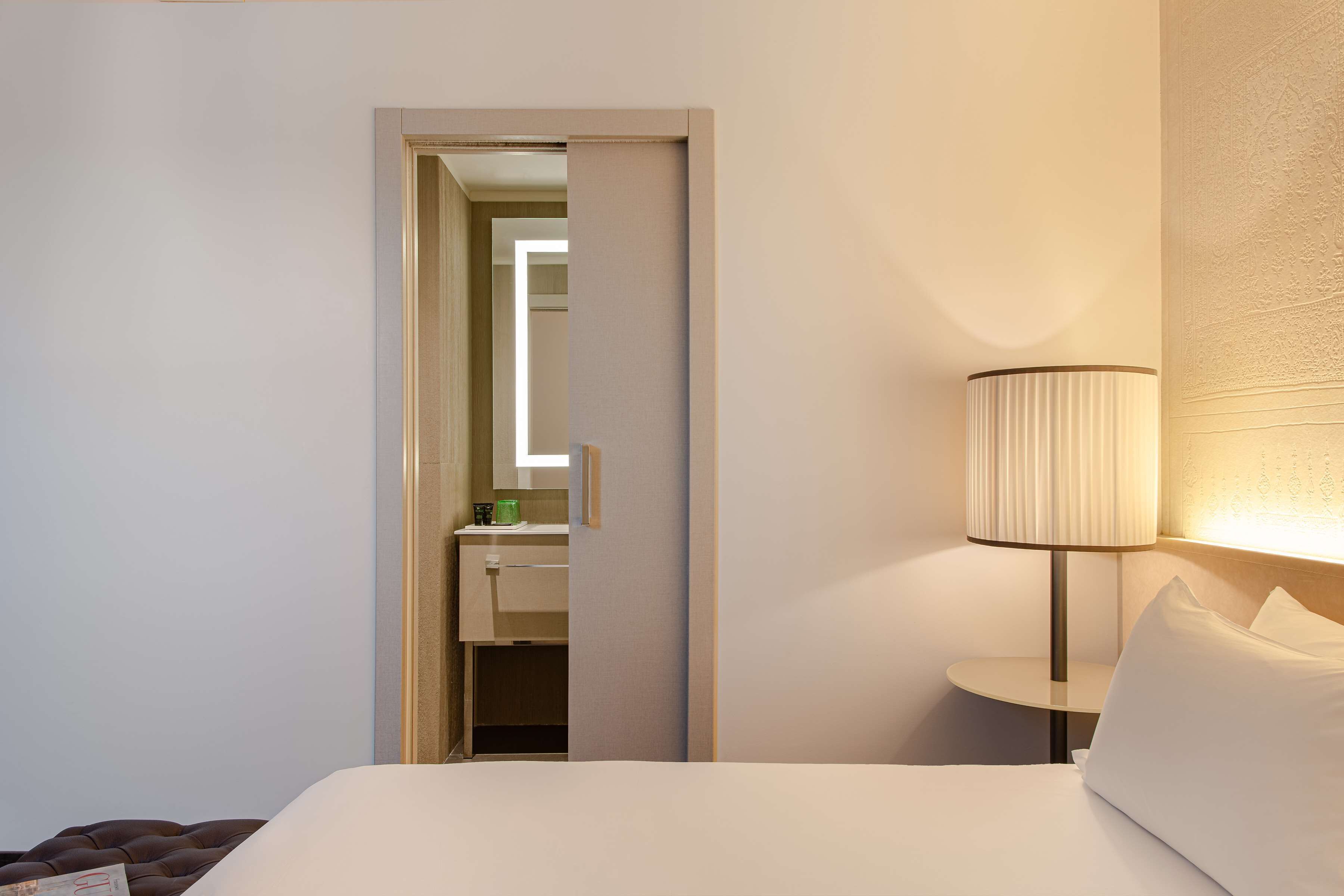 Quarto de hotel com uma cama, luminária de cabeceira e vista para o banheiro.