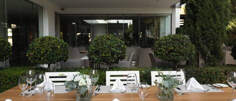 Mesa de jantar posta para refeição em ambiente externo, com arquitetura moderna, incluindo paredes de vidro e ventoinha de teto.