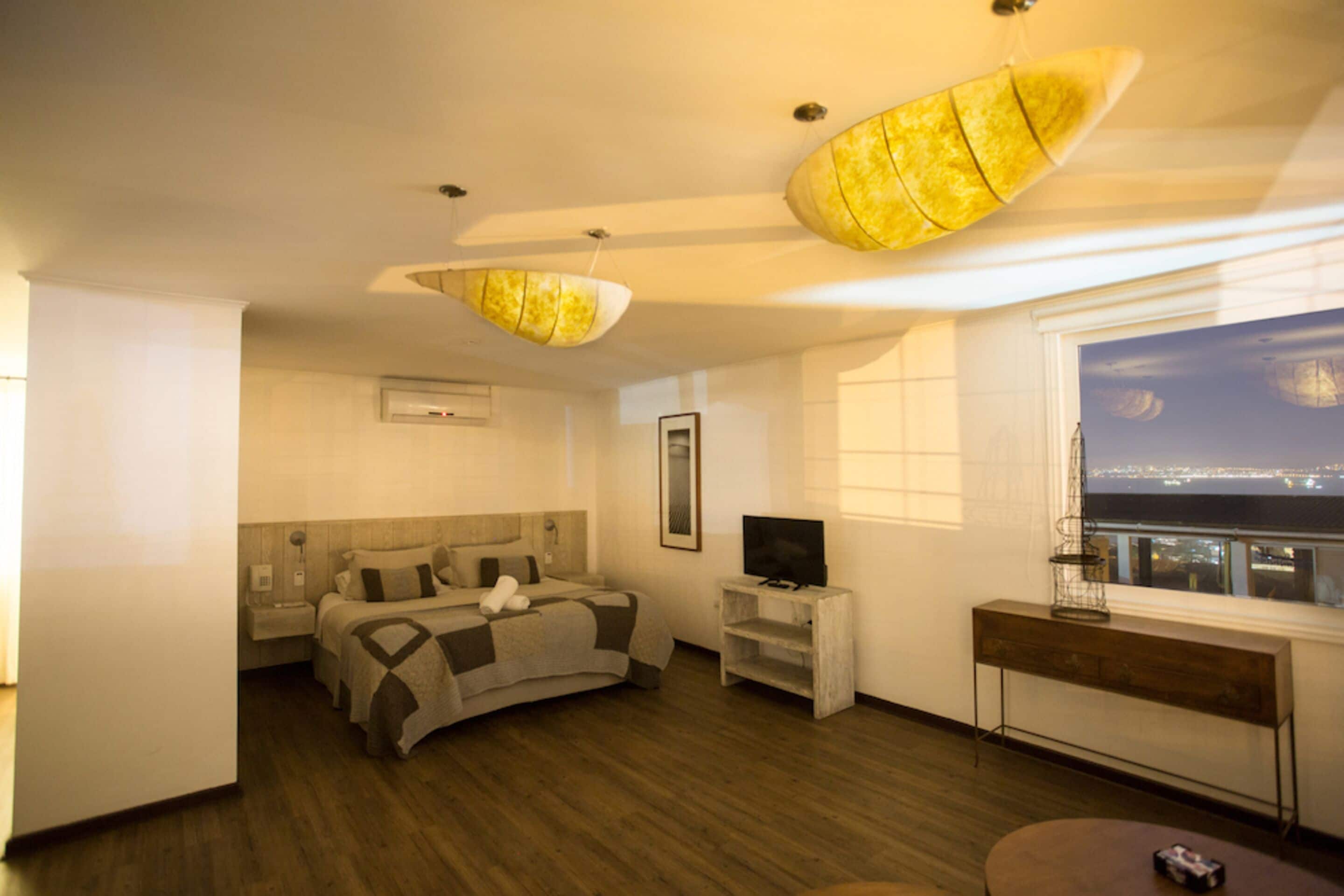 Quarto de hotel moderno com uma cama, uma televisão, uma escrivaninha e uma janela grande com vista para a cidade.