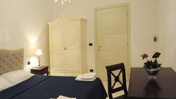 Quarto de hotel com cama, mesa de cabeceira com abajur, guarda-roupa, mesa de jantar e um vaso de flores, além de uma porta.