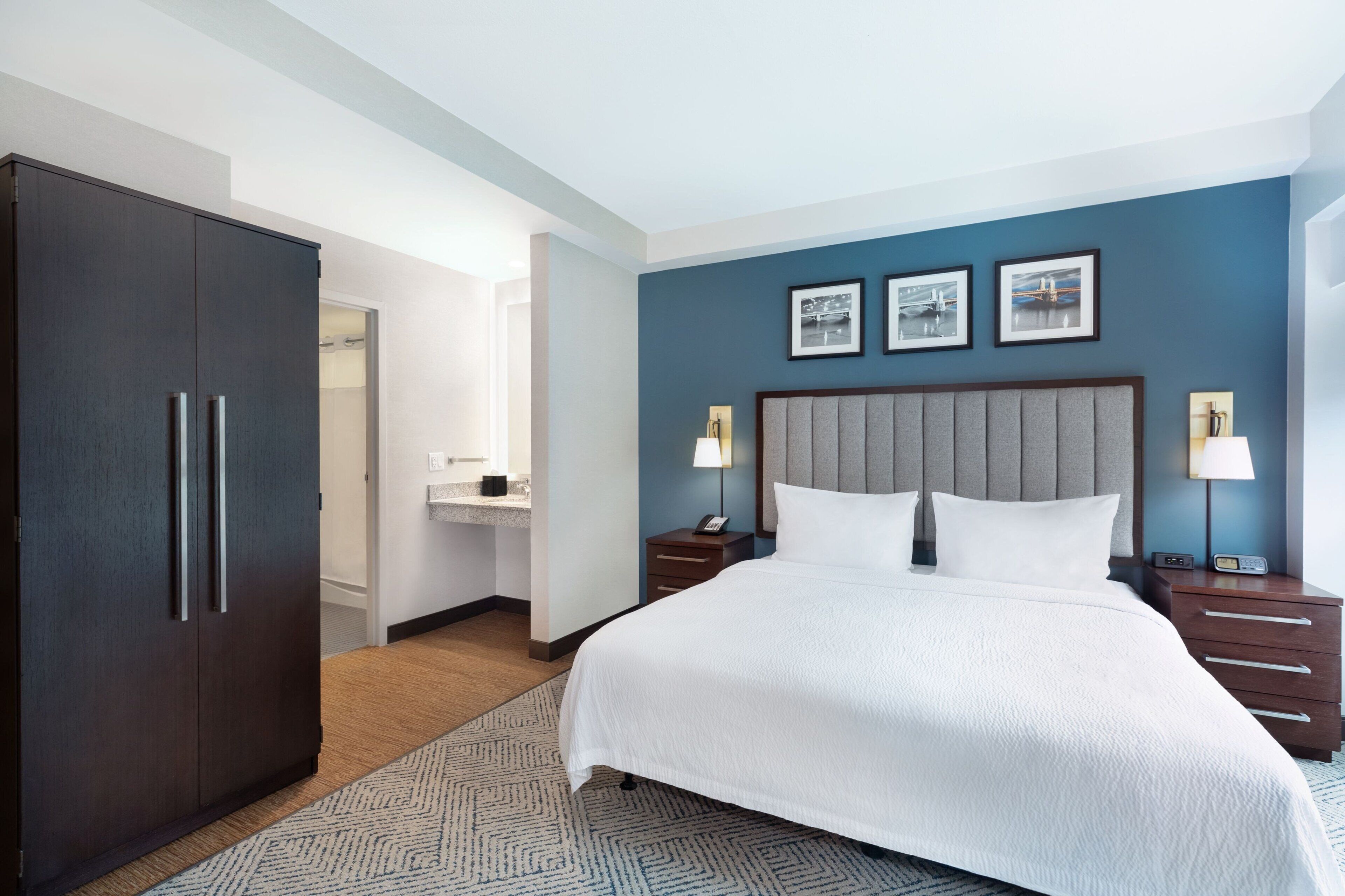 Quarto de hotel com uma cama grande, um guarda-roupa, uma toucador com espelho e três quadros na parede.