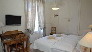 Quarto com cama, escrivaninha, cadeira, televisão e janela com cortinas.