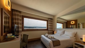 Quarto de hotel com uma cama grande, uma escrivaninha com um computador, uma cadeira, uma mesa de cabeceira e vista para um lago pela janela.
