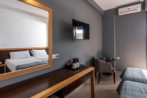 Quarto de hotel com cama, escrivaninha com espelho, televisão, cadeira e pequena mesa com um conjunto de objetos.