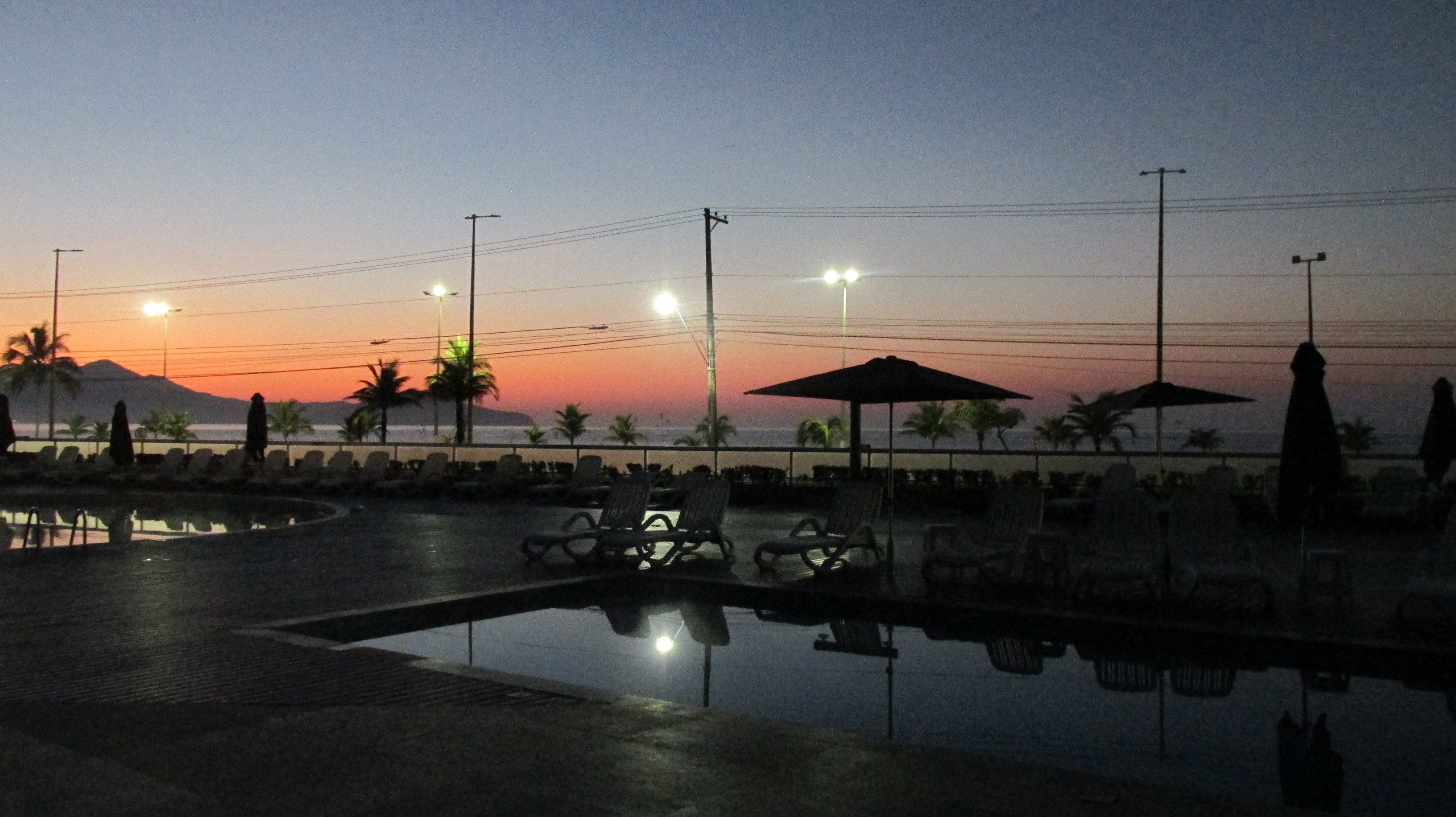 Área à beira da piscina com espreguiçadeiras e guarda-sóis, vista desobstruída do pôr do sol e superfície da água calma refletindo o entorno.