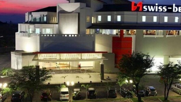 Um hotel moderno com um logotipo chamativo, vários andares e um estacionamento com diversos carros.