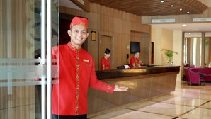 Área de recepção de hotel com recepcionista de uniforme vermelho, balcão de atendimento e espaço de espera com planta.