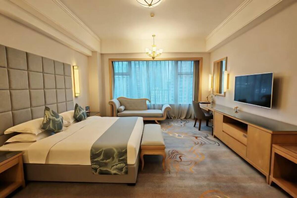 Quarto de hotel com uma cama grande, uma escrivaninha, uma televisão e uma janela com cortinas.