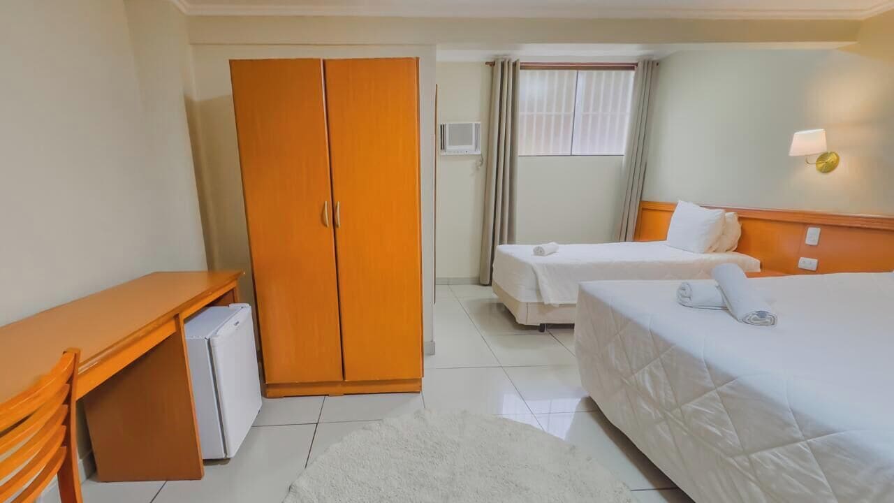 Quarto de hotel com duas camas, uma escrivaninha, um guarda-roupa e um pequeno frigorífico.