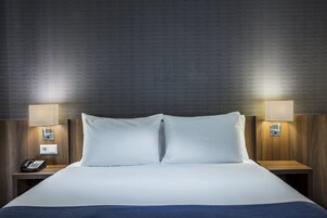 Quarto de hotel com uma cama grande, duas mesas de cabeceira, um telefone e luminárias fixadas na parede.