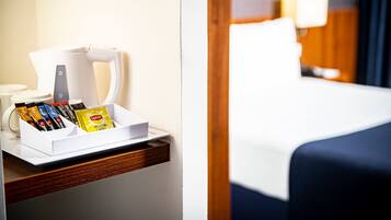 Quarto de hotel com cama bem arrumada, mesa de cabeceira com cafeteira e bandeja de petiscos.