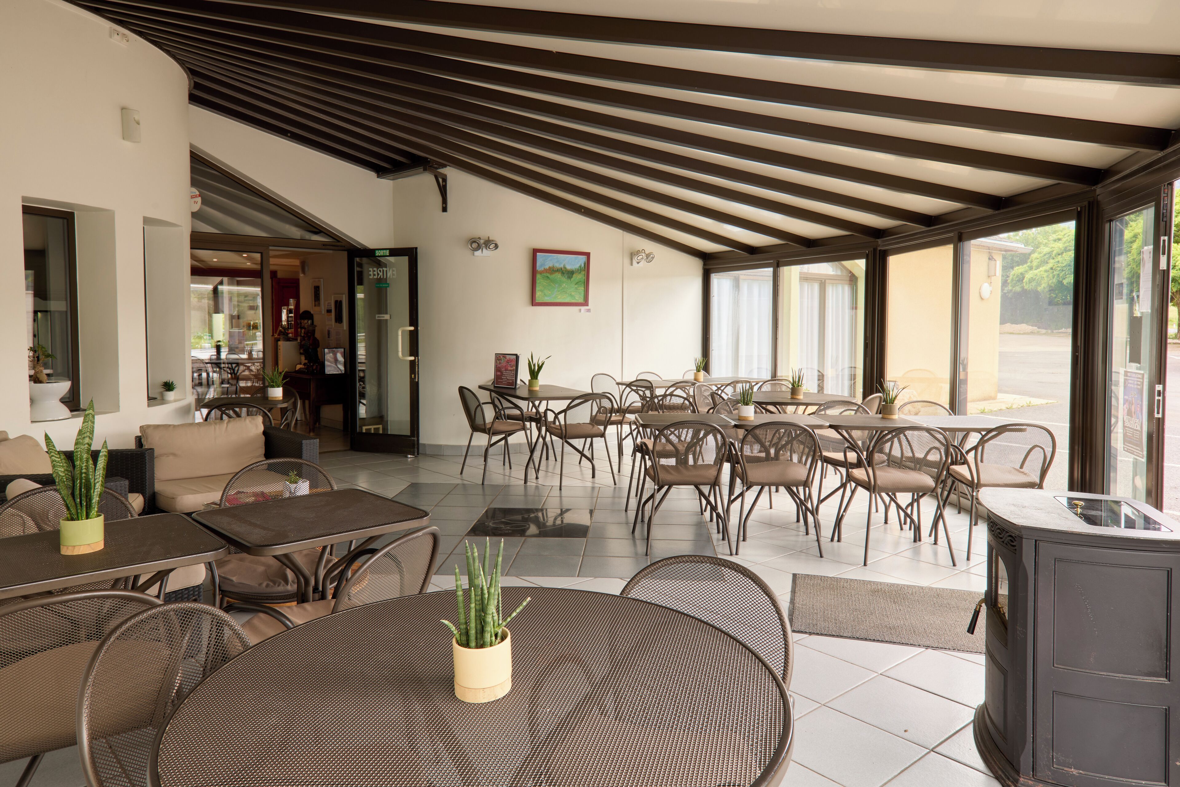 Um restaurante com mesas e cadeiras ao ar livre, incluindo mesas redondas, e uma grande janela que oferece vista para a rua.
