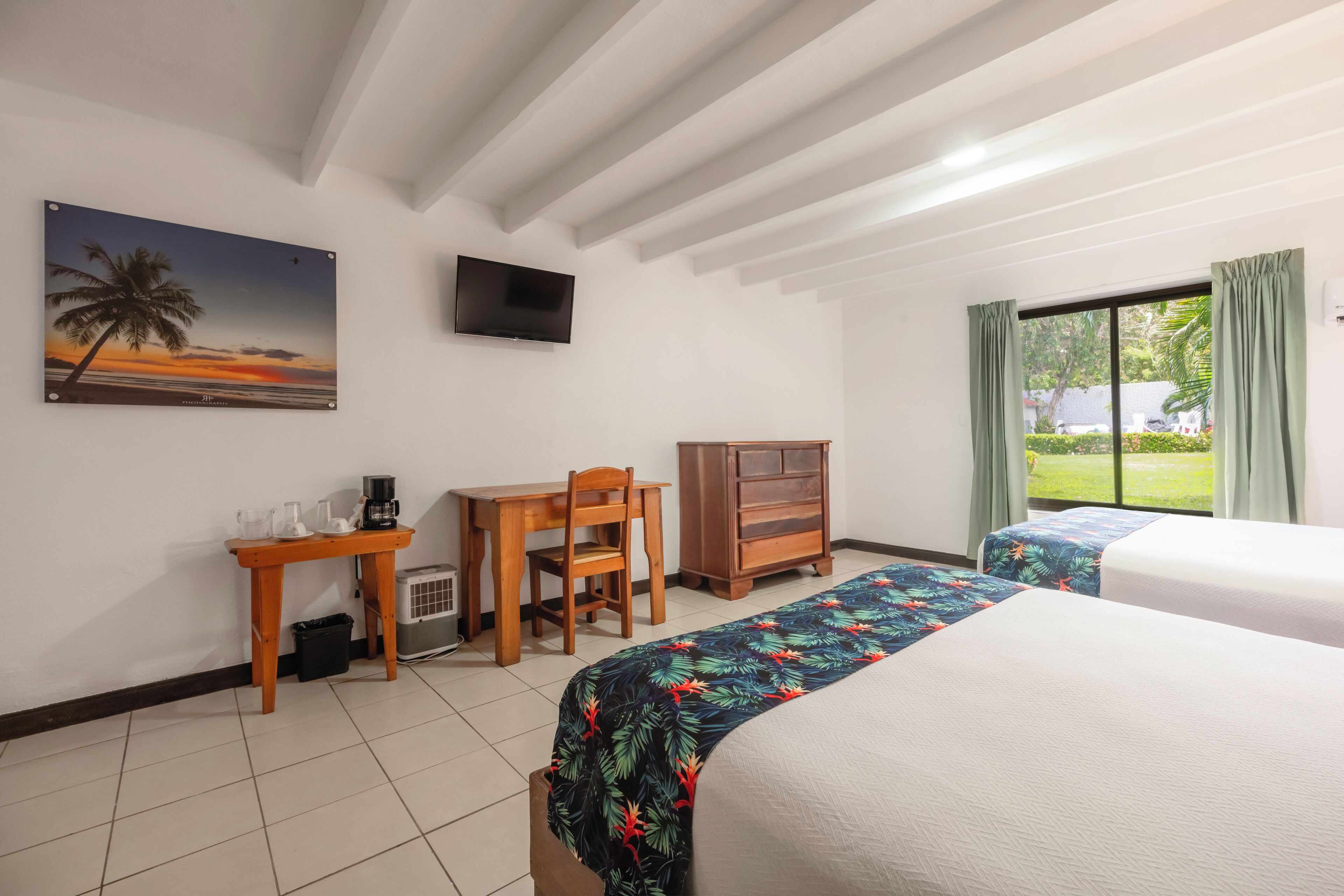 Quarto de hotel com duas camas, uma escrivaninha com cadeira, máquina de café, televisão na parede e um quadro de um pôr do sol.