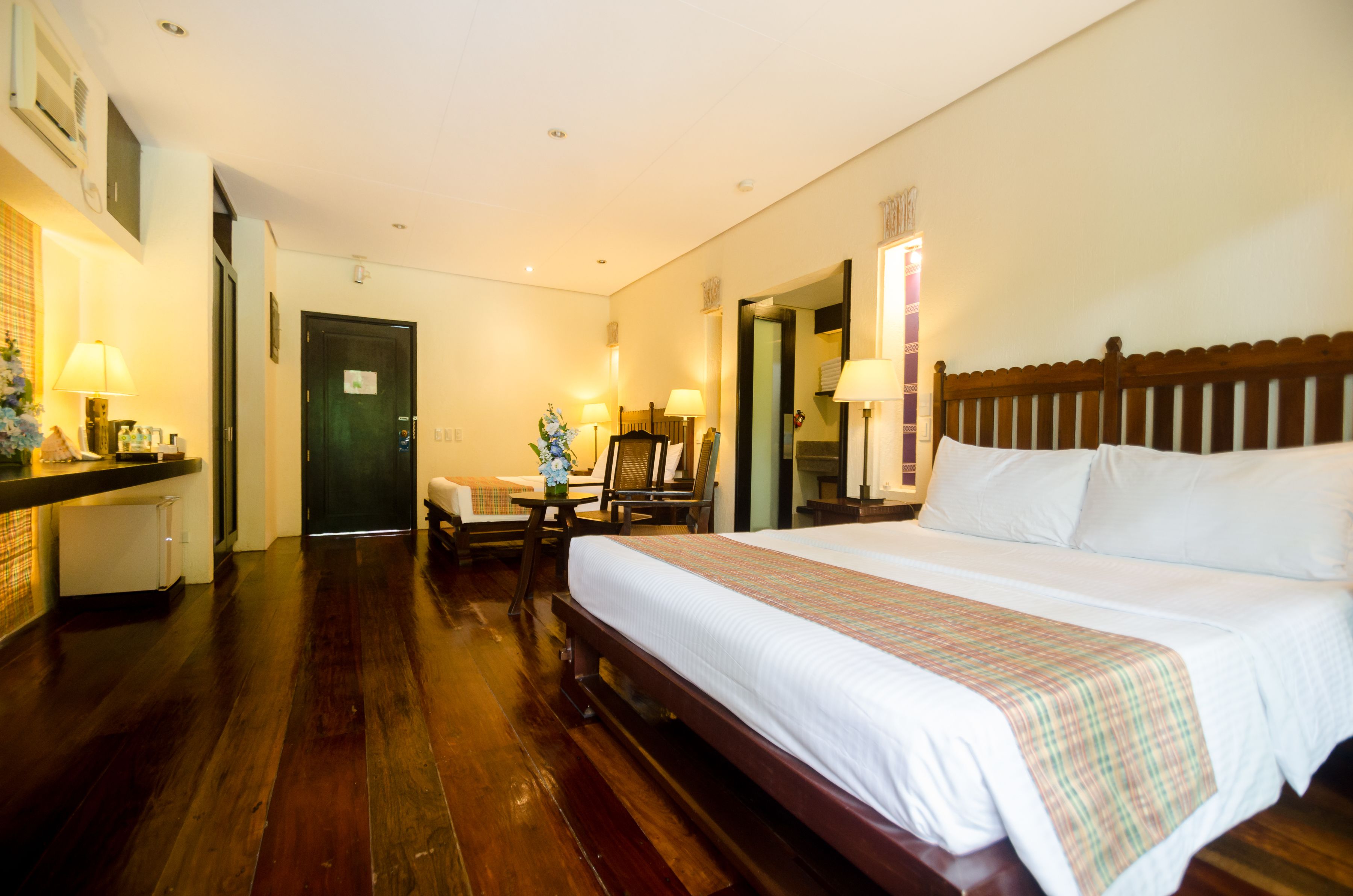 Quarto de hotel com uma cama grande, piso de madeira, uma escrivaninha com abajur e uma cadeira.
