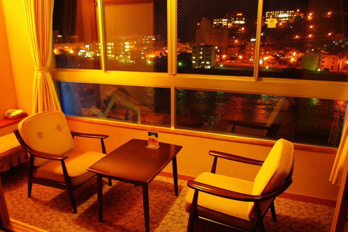 Quarto com duas cadeiras, uma pequena mesa, uma janela com vista para as luzes da cidade e uma cortina à esquerda.