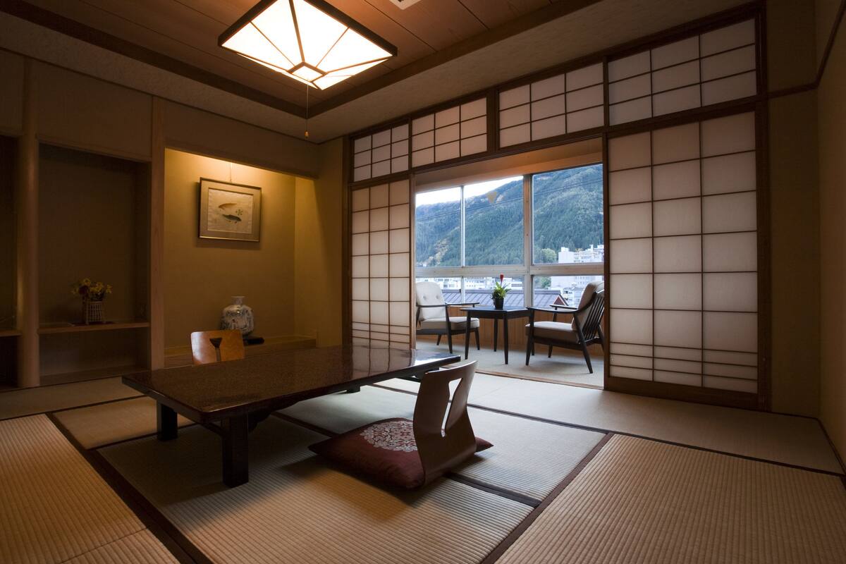 Um quarto japonês tradicional com tatames, uma mesa baixa, um banco almofadado, um quadro emoldurado e vista para uma montanha através de biombos shoji.