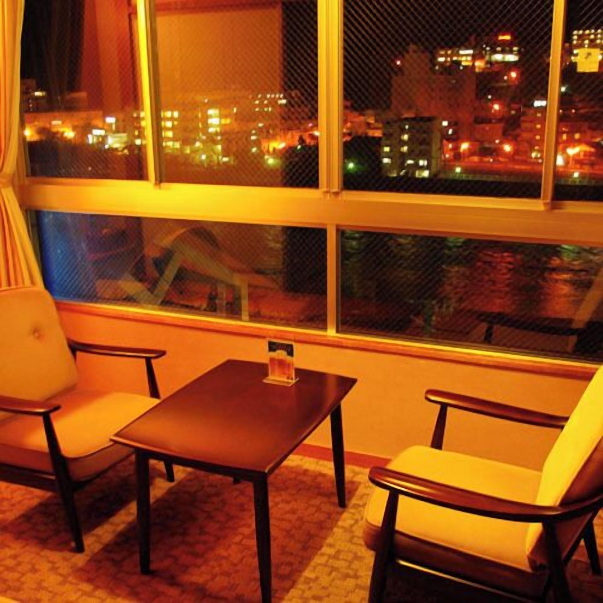 Quarto com uma janela grande, duas cadeiras, uma mesa pequena e vista para a cidade iluminada à noite.