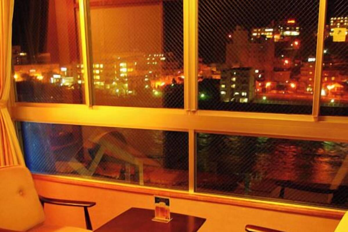 Quarto com uma janela grande, duas cadeiras, uma mesa pequena e vista para a cidade iluminada à noite.