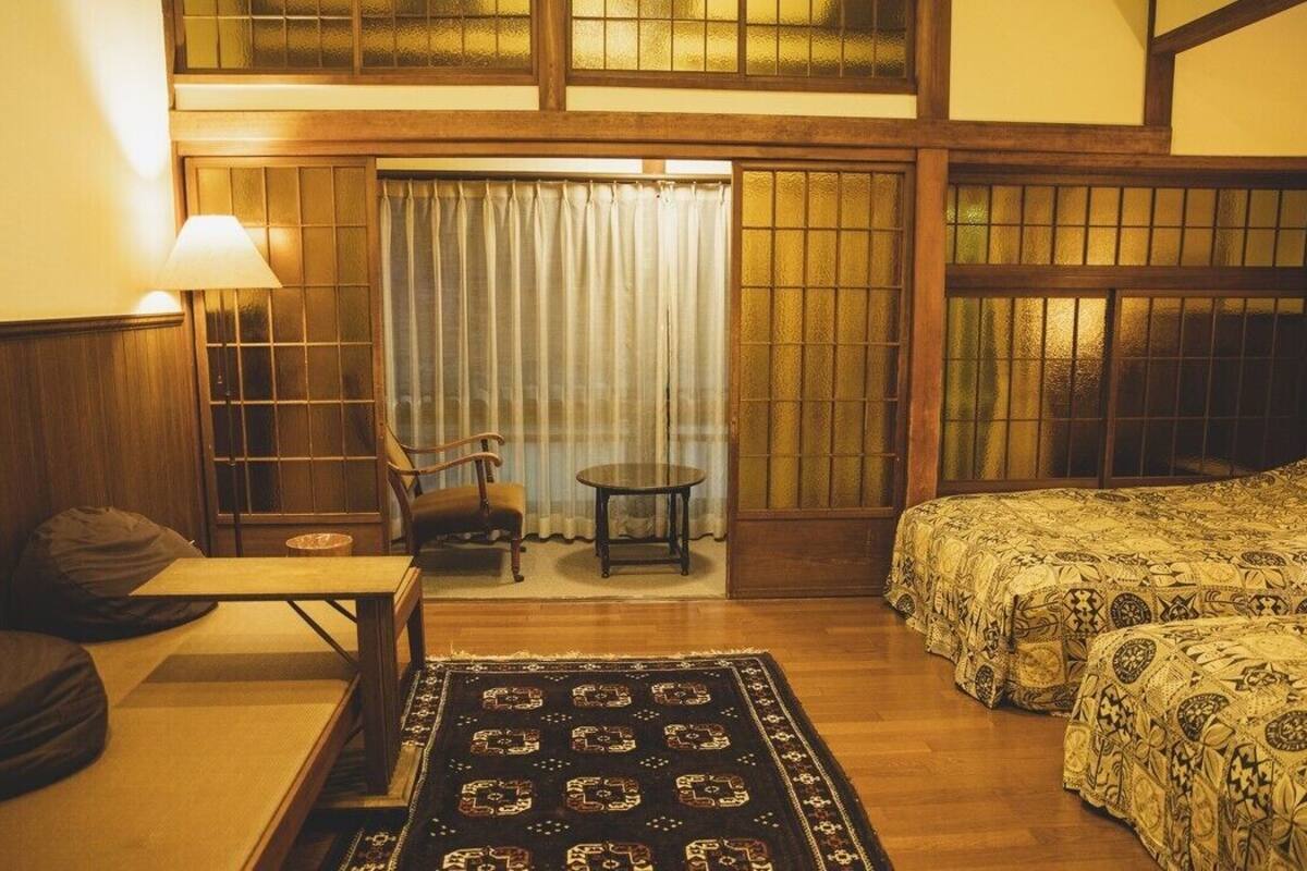 Um quarto tradicional japonês com piso de tatame, uma mesa baixa com almofadas, futon e uma pequena mesa redonda, além de uma porta corrediça com cortina.