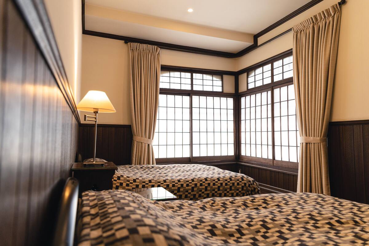 Quarto de hotel com duas camas, uma mesa de cabeceira, uma luminária e janelas amplas com cortinas.