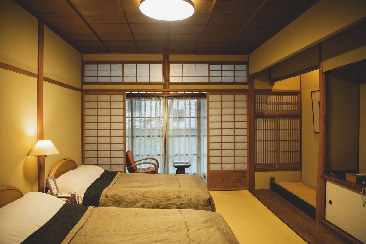 Quarto japonês tradicional com tatames, duas camas, uma escrivaninha de madeira e uma porta de correr.