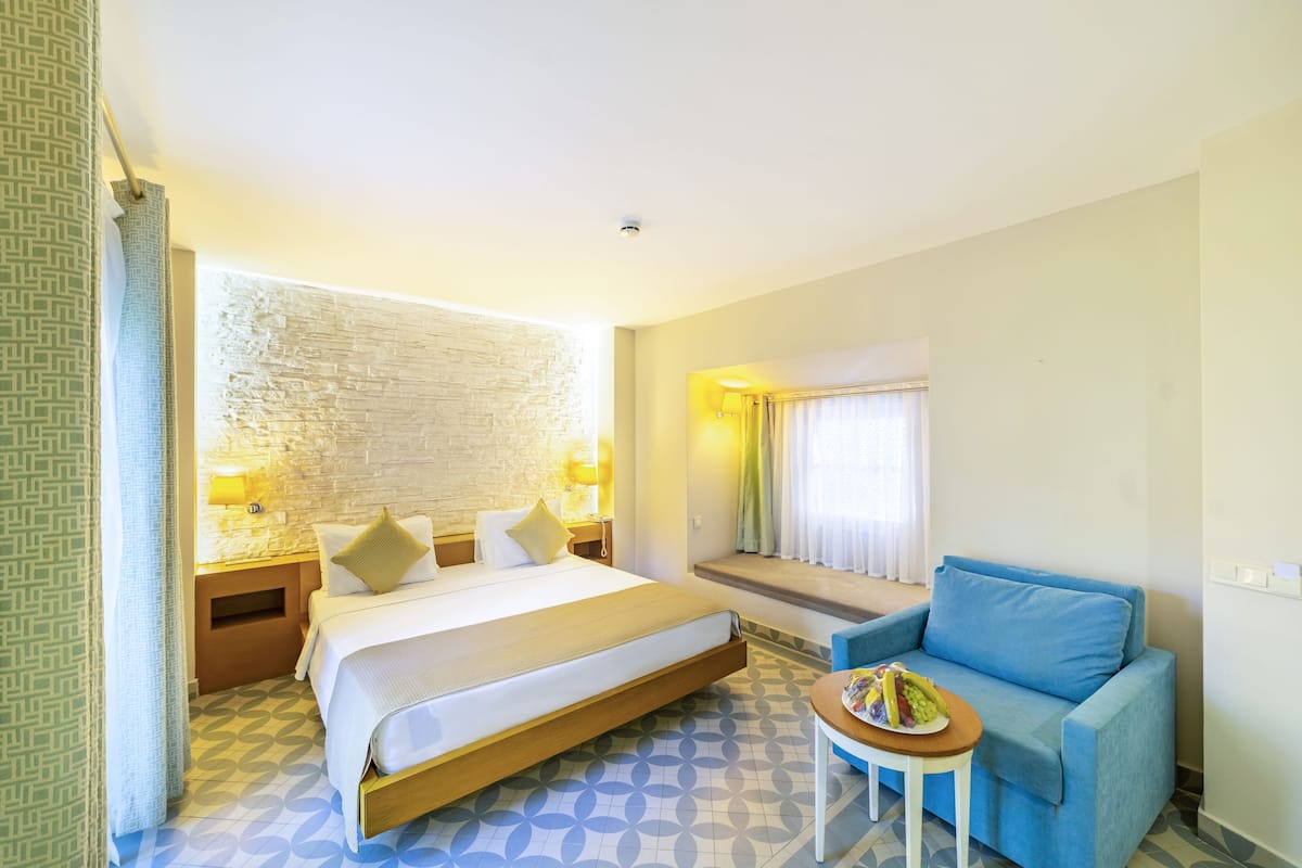 Quarto de hotel com uma cama grande, um sofá azul, uma mesinha com frutas e uma janela com cortinas.