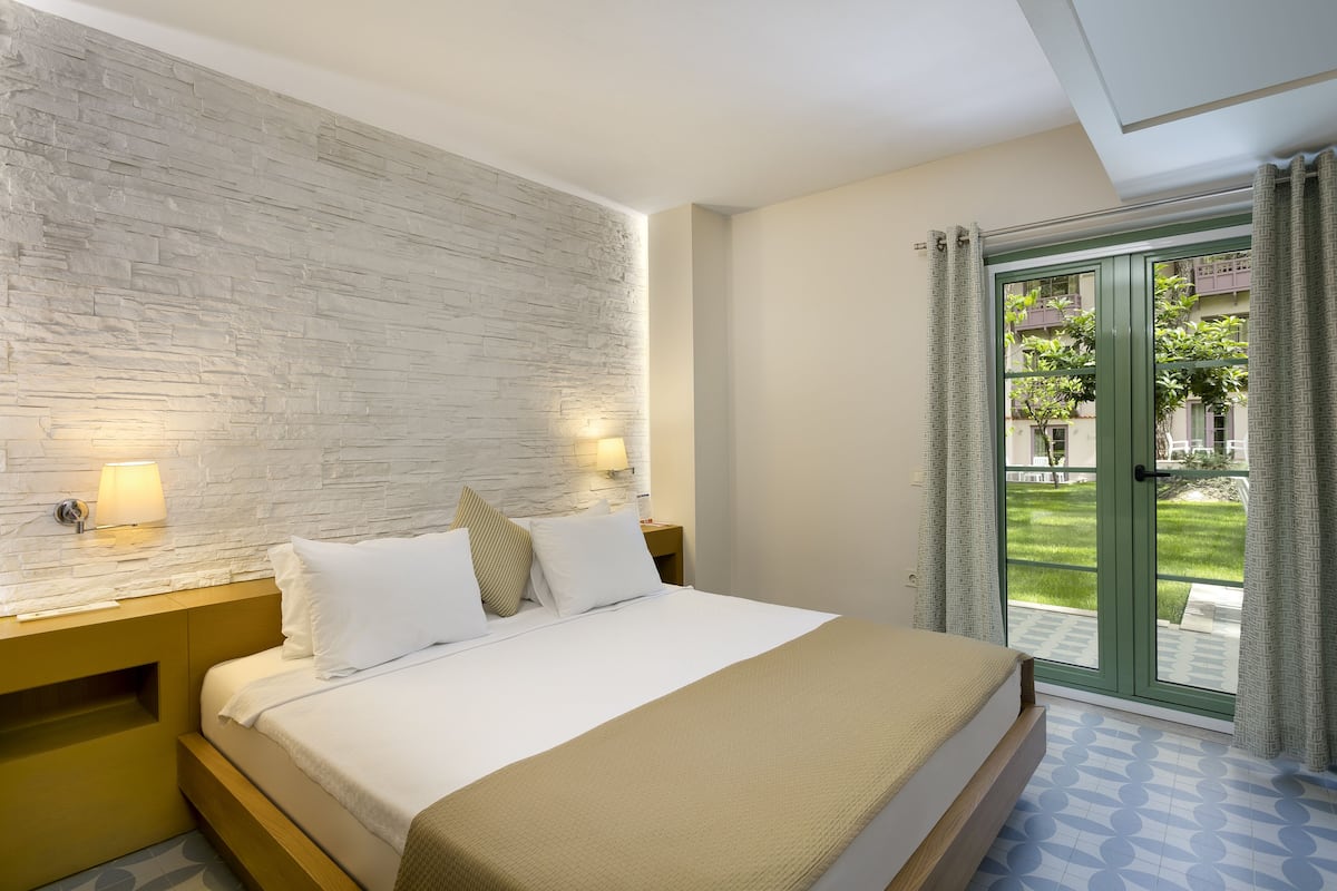 Quarto de hotel moderno com parede texturizada em pedra, cama com roupa de cama branca e vista através de uma porta de vidro para uma área verde externa.