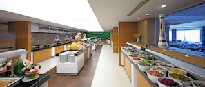 Buffet com diversas estações de comida e área de cozinha ao fundo.
