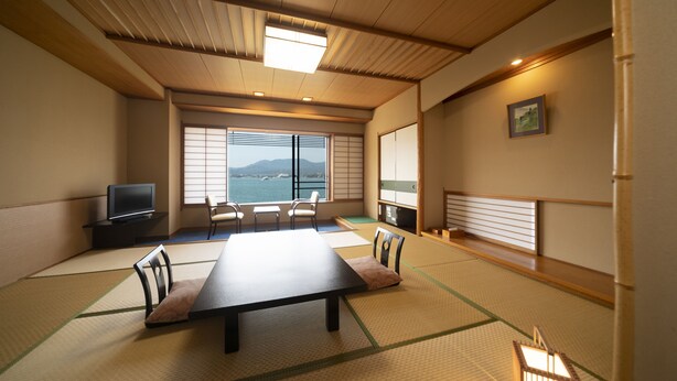 Um quarto japonês tradicional com tatames, uma mesa baixa, cadeiras, televisão e vista para o mar através de grandes janelas.