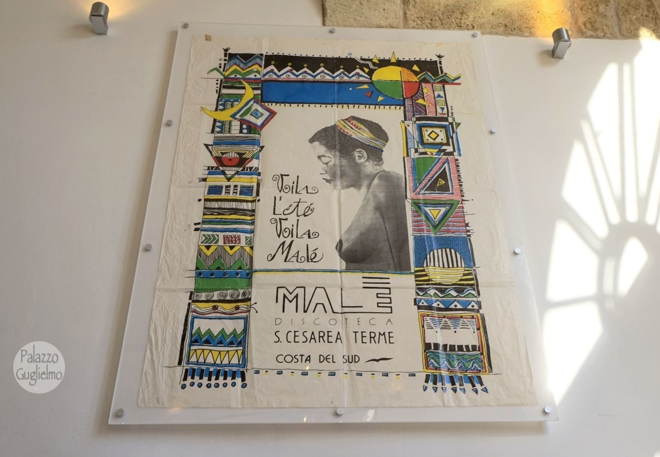 Uma obra de arte emoldurada com uma figura central e padrões decorativos, fixada em uma parede.