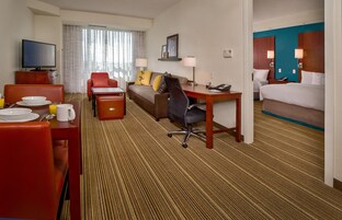 Quarto de hotel com cama, escrivaninha, cadeira, televisão e área de jantar com mesa e cadeiras.
