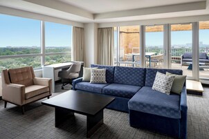 Quarto de hotel moderno com uma janela grande, um sofá, uma cadeira, uma mesa de trabalho e uma varanda com mobiliário exterior.
