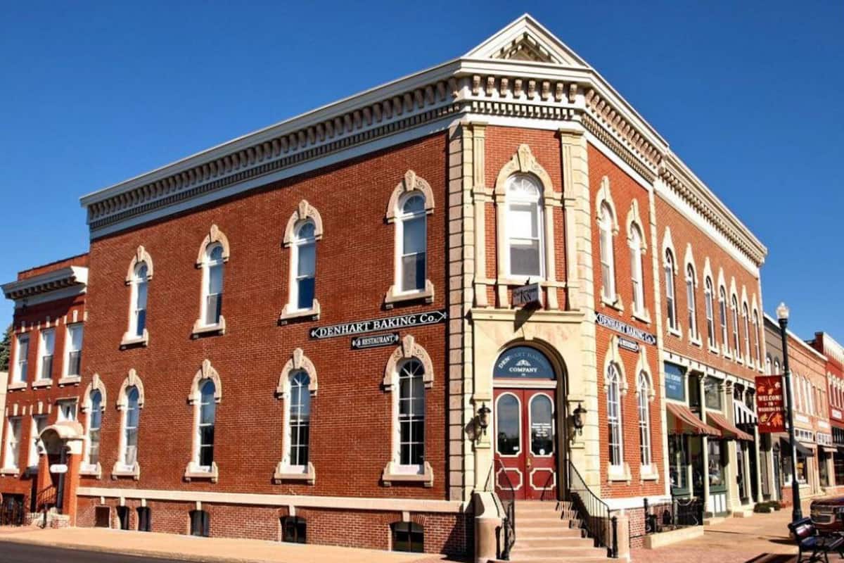 Um edifício histórico de tijolos vermelhos com entrada na esquina, exibindo uma placa da Denhart Baking Co. e uma escadaria visível que leva à entrada.