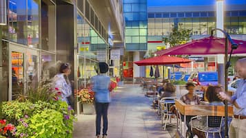 Café ao ar livre com mesas e cadeiras, rodeado por edifícios de vidro e um guarda-sol vermelho.