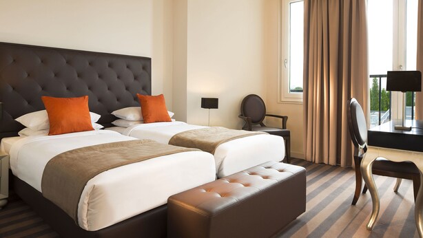Quarto de hotel com duas camas, uma cabeceira grande com detalhes em capitonê, um banquinho, uma escrivaninha com cadeira e vista para o exterior através de uma janela com cortinas.