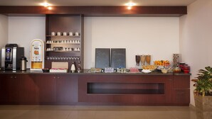 Uma área de recepção com uma máquina de café, uma mesa de frutas e um quadro negro com escrita.