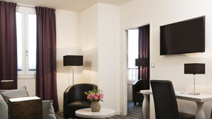 Quarto de hotel com sofá, uma mesa de jantar com duas cadeiras, televisão na parede e um vaso com flores sobre uma mesinha.